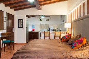 Quarto com cama grande, escrivaninha e uma kitchenette ao fundo.