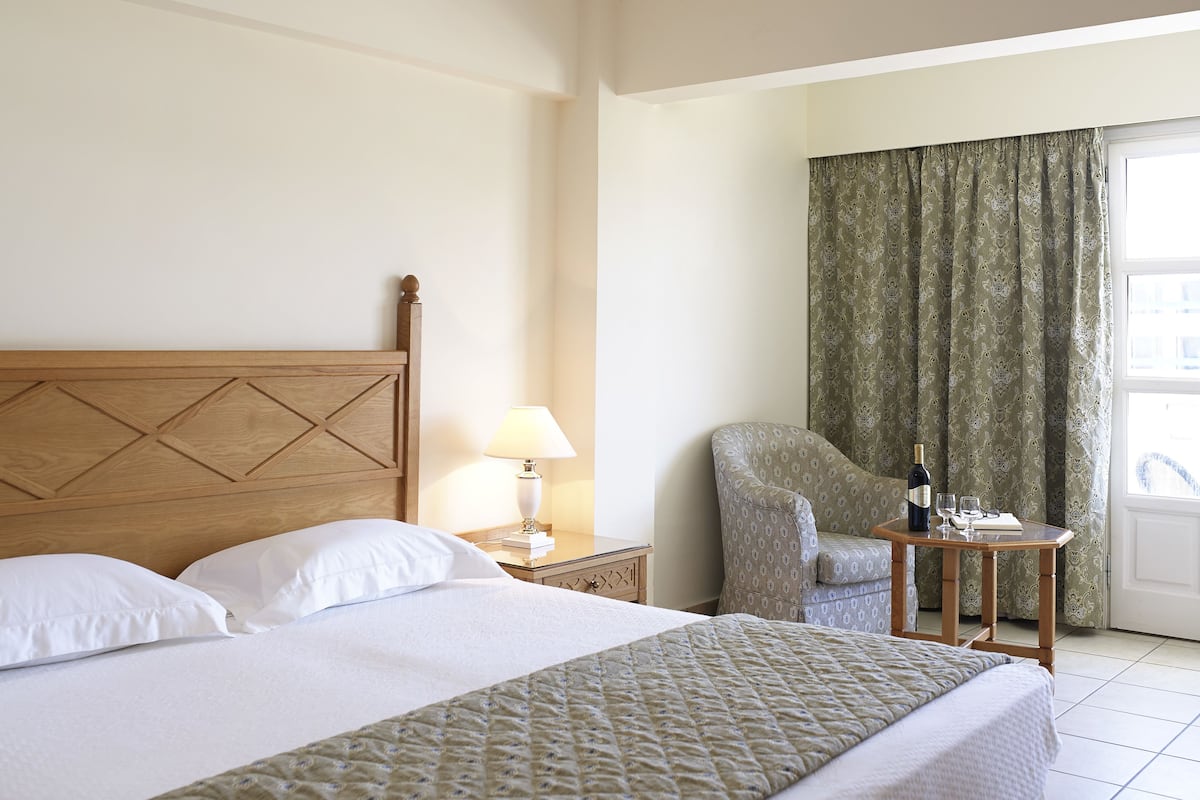 Quarto de hotel com cama, cadeira, mesa de cabeceira com abajur, pequena mesa com garrafa e copos, e janela com cortinas.
