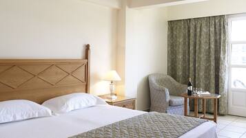 Quarto de hotel com cama, cadeira, mesa de cabeceira com abajur, pequena mesa com garrafa e copos, e janela com cortinas.