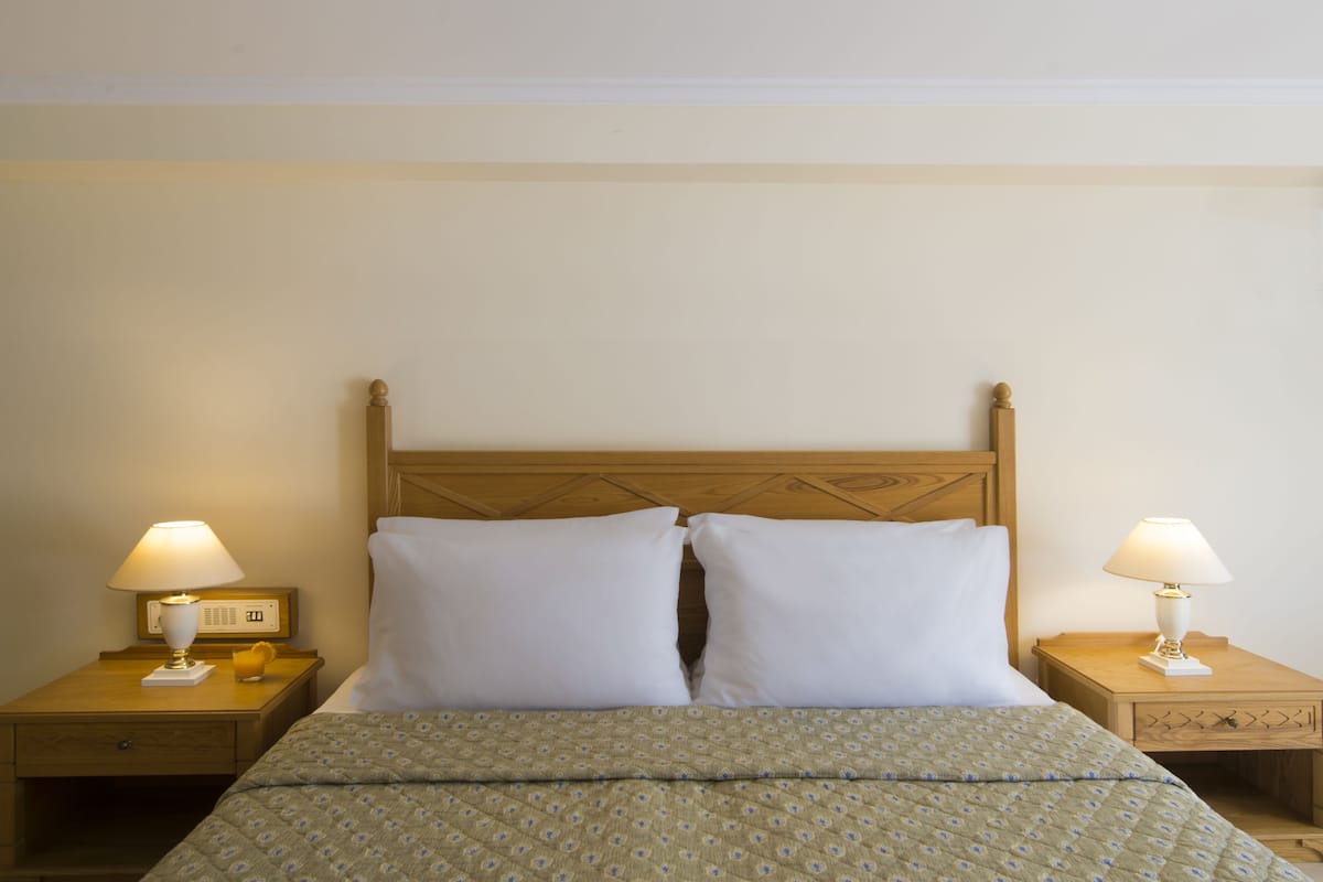 Uma cama bem arrumada com almofadas brancas e um edredom estampado, com duas mesas de cabeceira de cada lado, com luminárias e um rádio.