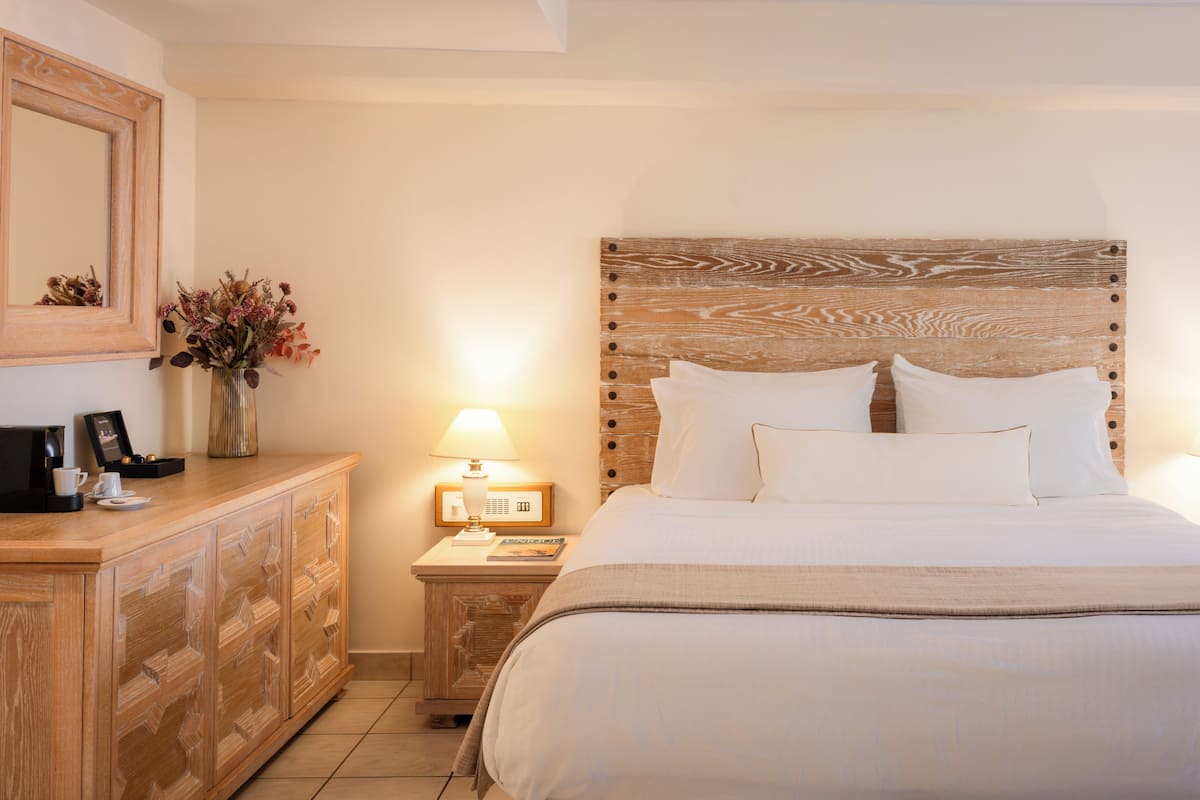 Quarto de hotel bem arrumado, com cama, abajures, cabeceira de madeira, cômoda e um vaso com flores secas.