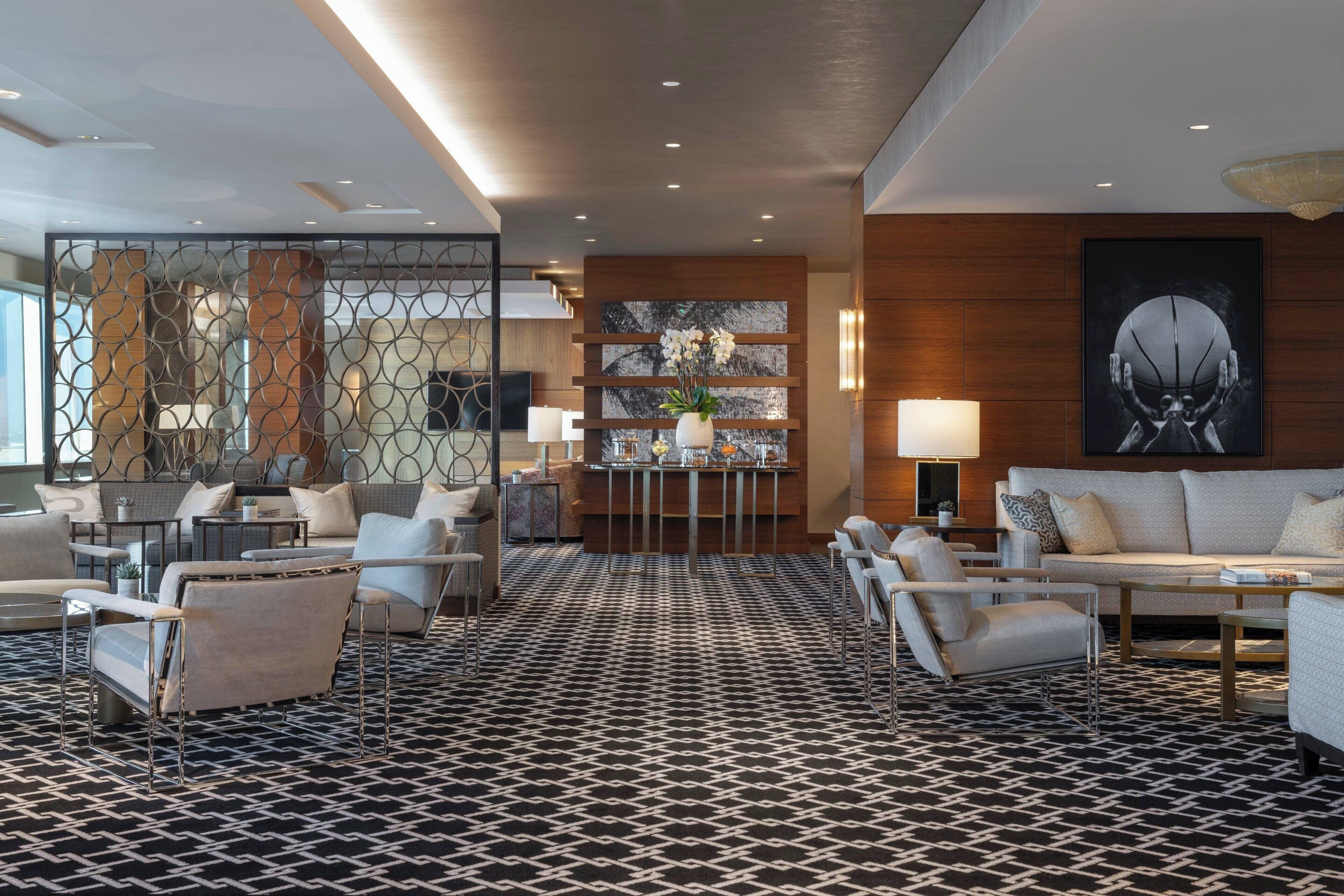 Um hall de hotel moderno com um piso de padrão geométrico, uma área de estar central com sofás e cadeiras, uma parede decorativa com uma obra de arte com tema de basquete e uma televisão fixada na parede.