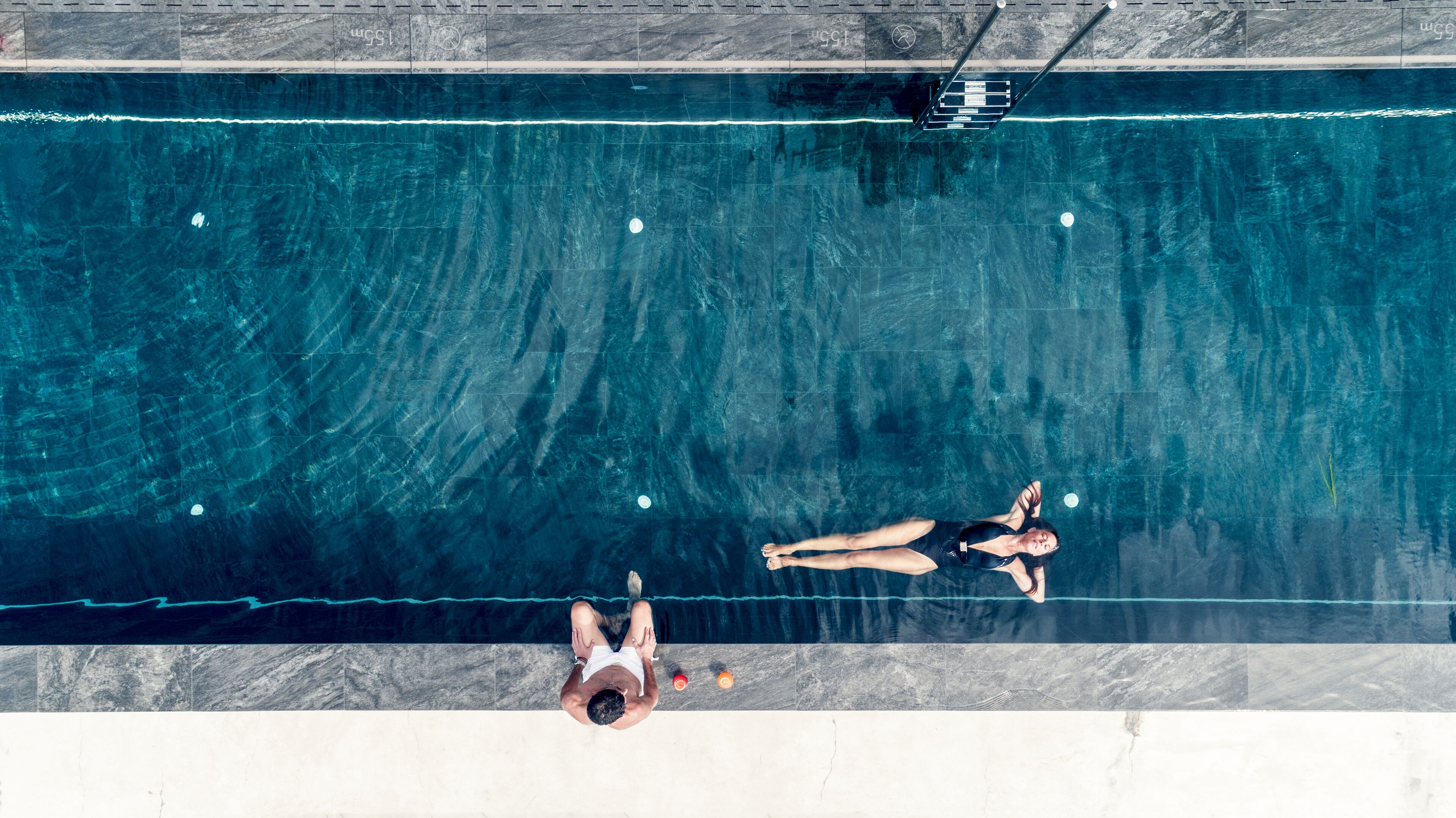 Vista aérea de uma piscina com duas pessoas nadando e uma terceira à beira da piscina.