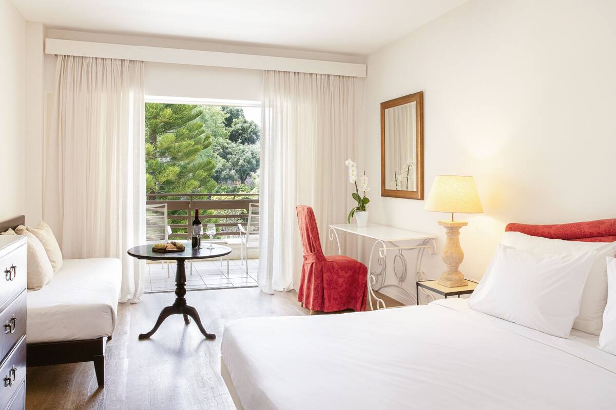 Quarto de hotel com uma cama, uma escrivaninha, uma cadeira, uma mesinha com uma garrafa e copos, e vista para o verde lá fora.