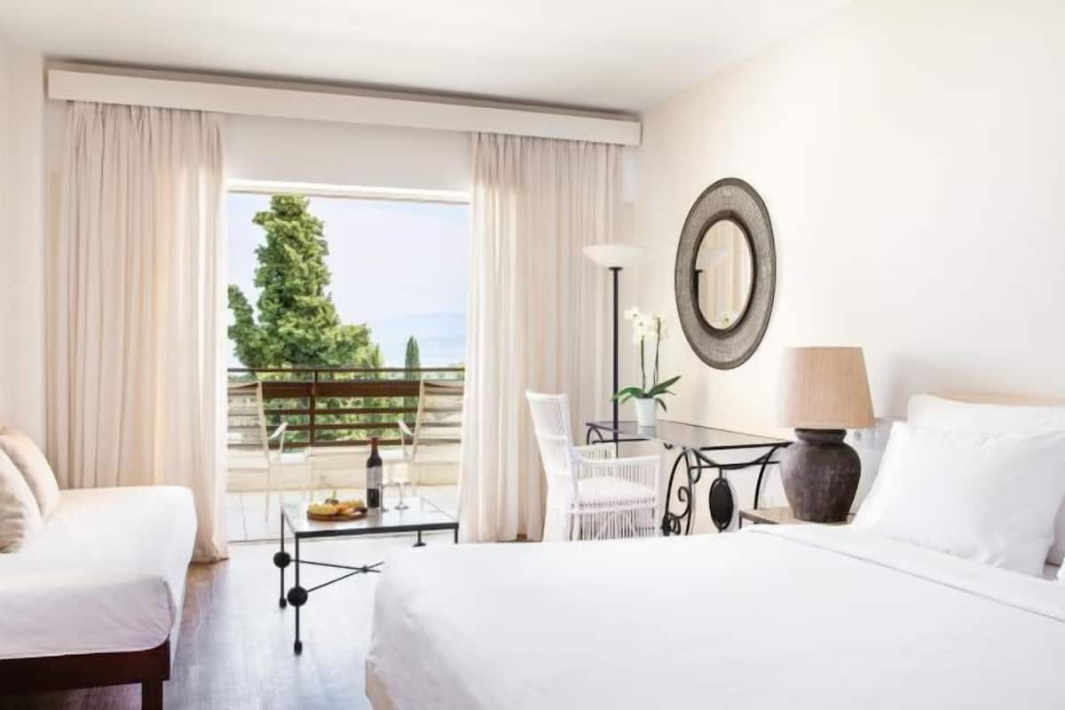 Quarto de hotel com cama, escrivaninha, cadeira, luminária e vista para o verde lá fora.