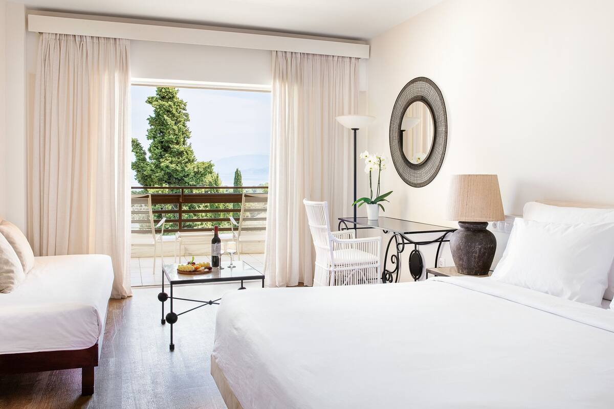 Quarto de hotel com cama, escrivaninha e cadeira, mesa com garrafa e copos, luminária, e vista para árvores e montanhas.