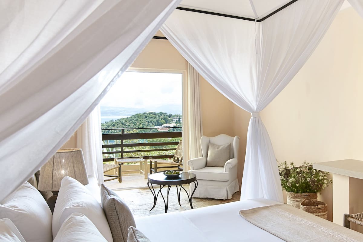 Quarto com cama, uma cadeira, uma mesinha e varanda com vista para o verde.