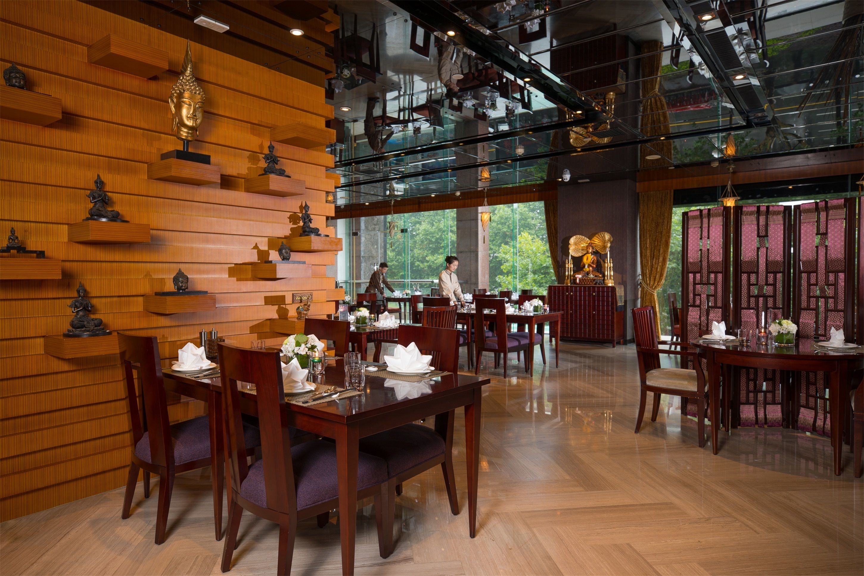 Restaurante com painéis de madeira, teto de vidro e várias estátuas de Buda. Mesas dispostas com toalhas brancas e arranjos florais.