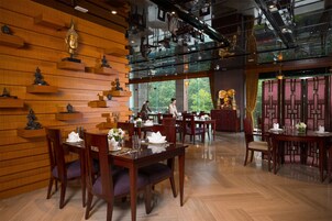 Restaurante com painéis de madeira, teto de vidro e várias estátuas de Buda. Mesas dispostas com toalhas brancas e arranjos florais.