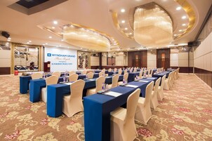 Uma sala de conferências com fileiras de mesas, toalhas de mesa azuis e cadeiras bege. Uma tela exibe o logotipo Wyndham Grand.