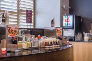 Balcão de café da manhã com opções de waffle, dispensadores de xarope e uma tela de TV exibindo os itens do cardápio.