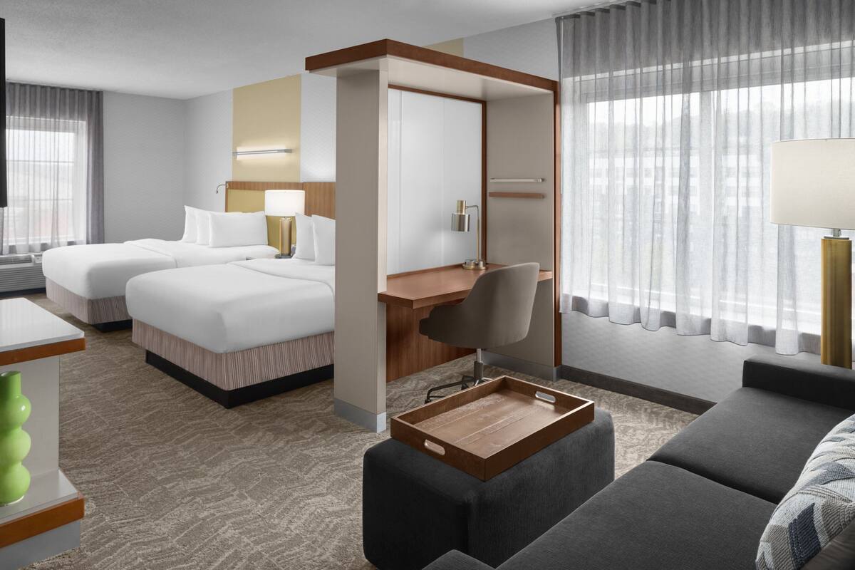 Quarto de hotel com duas camas, uma escrivaninha, uma cadeira, um sofá e uma mesinha de apoio.