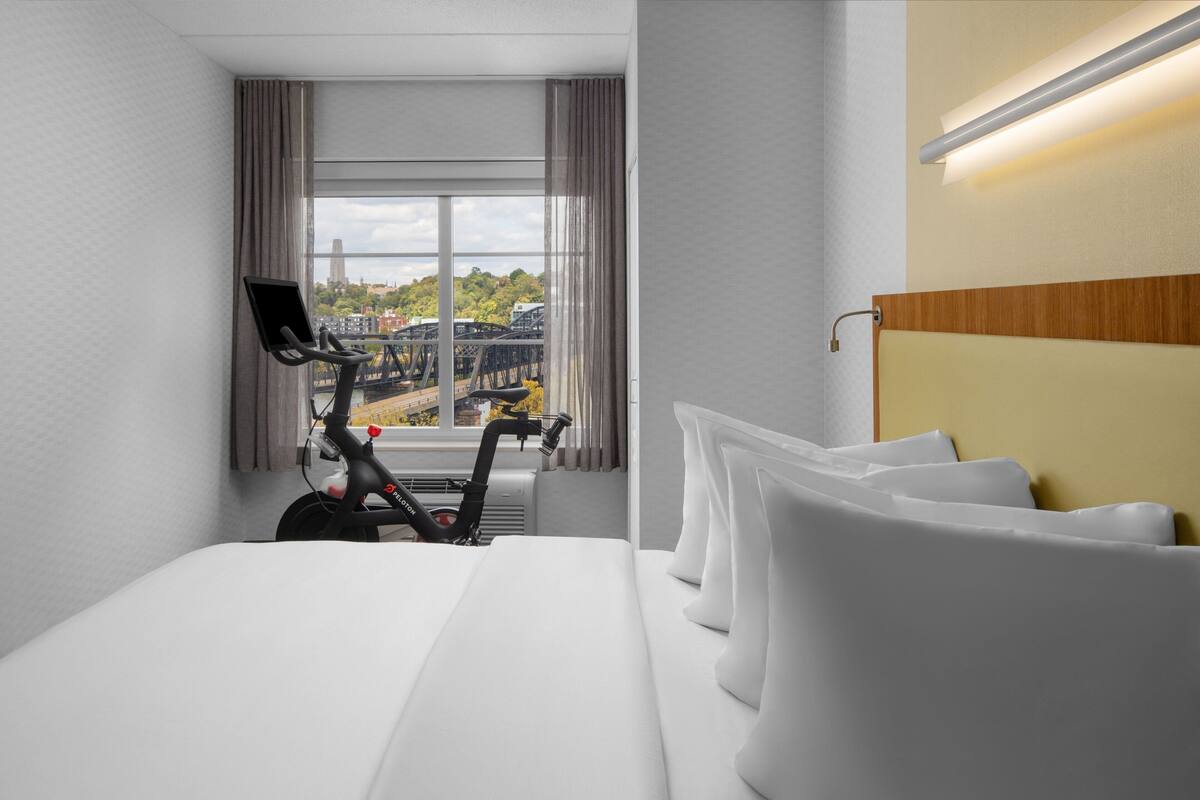 Quarto de hotel com cama, bicicleta ergométrica e janela com vista para a cidade.