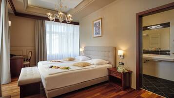 Quarto de hotel com uma cama grande, uma escrivaninha, uma cadeira, uma mesinha com flores, e um banheiro visível por uma porta aberta.