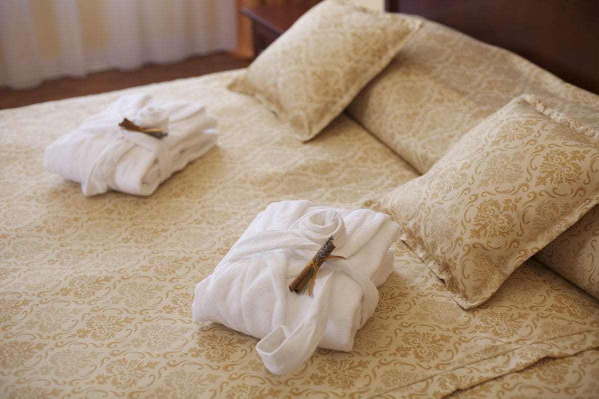 Cama bem arrumada com colcha estampada e dois travesseiros, cada um com uma toalha branca dobrada e um pequeno objeto decorativo.