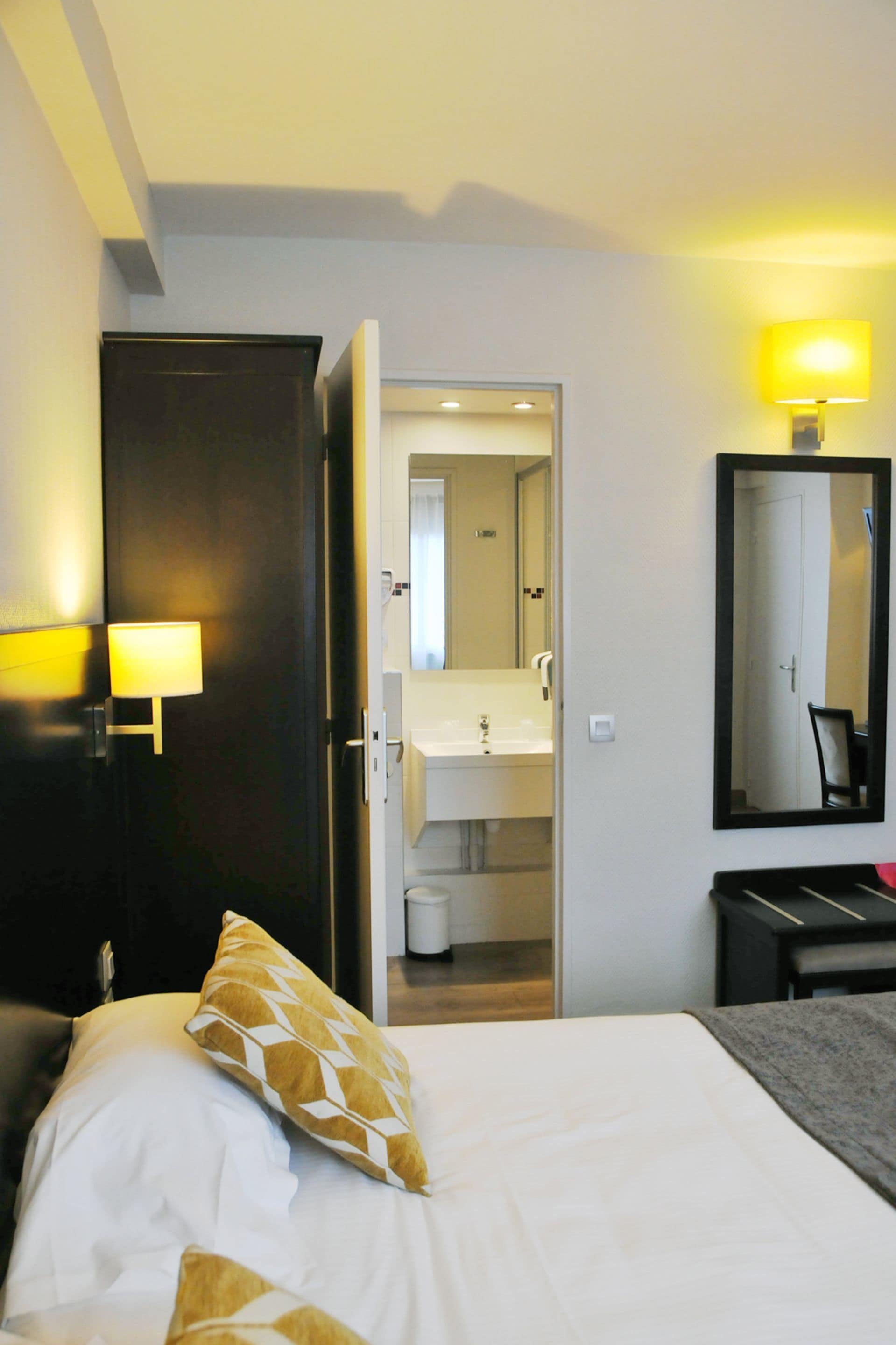 Quarto de hotel com cama, mesa de cabeceira, escrivaninha, espelho, banheiro com pia e espelho, e porta que dá para outro quarto.