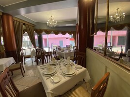 Uma sala de jantar com mesas preparadas para quatro pessoas, cadeiras de madeira e um lustre.