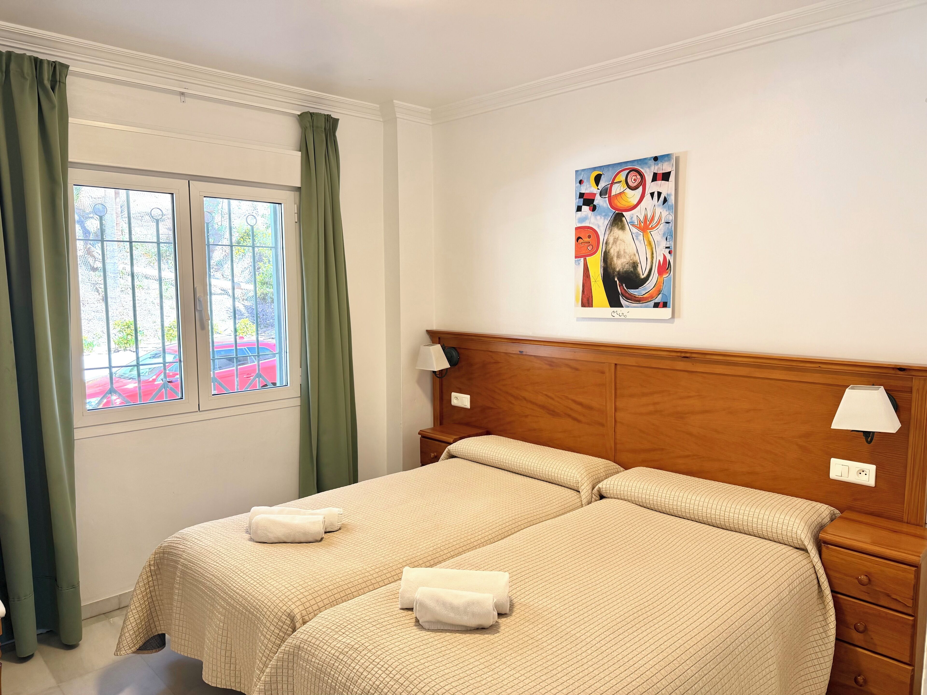 Quarto de hotel com duas camas, uma janela com cortinas, um quadro na parede e uma cabeceira de madeira.