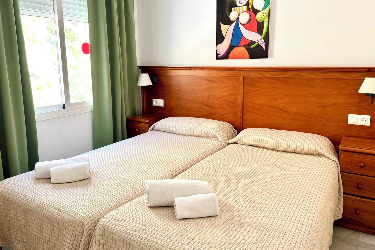 Quarto de hotel com duas camas, cabeceira de madeira, um quadro na parede e uma janela com cortinas.
