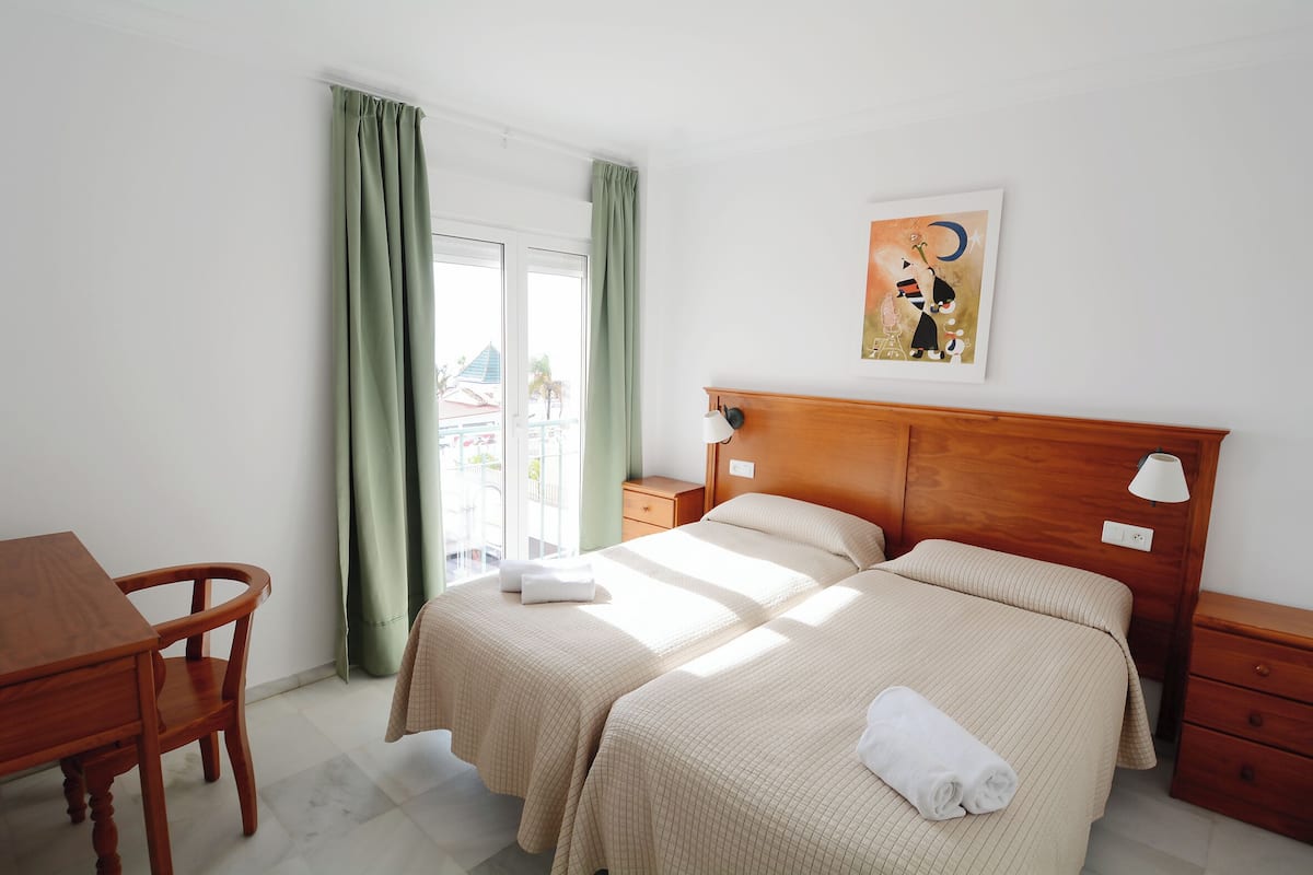 Quarto de hotel com duas camas, uma escrivaninha de madeira, uma mesa de cabeceira e uma janela com cortinas.