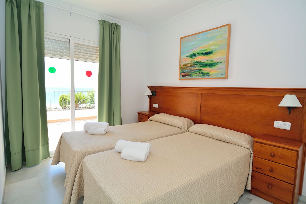 Quarto de hotel com duas camas, uma cabeceira de madeira, um quadro e vista para o mar.
