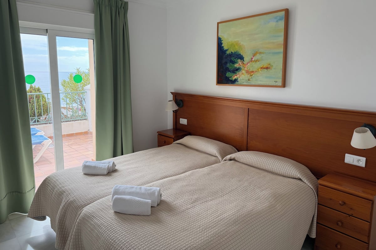 Quarto de hotel com cama, mesa de cabeceira, um quadro e vista para o mar através de uma porta deslizante.