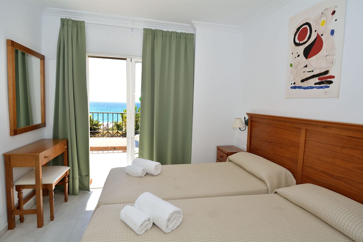 Quarto de hotel com cama, escrivaninha, cadeira e vista para o mar pela janela.