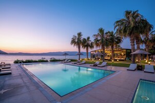 Uma área de piscina com espreguiçadeiras e guarda-sóis, um edifício iluminado e vista para o mar ao entardecer.
