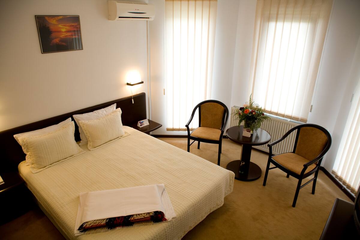 Quarto de hotel com uma cama grande, duas cadeiras, uma mesinha com um vaso e um quadro na parede.
