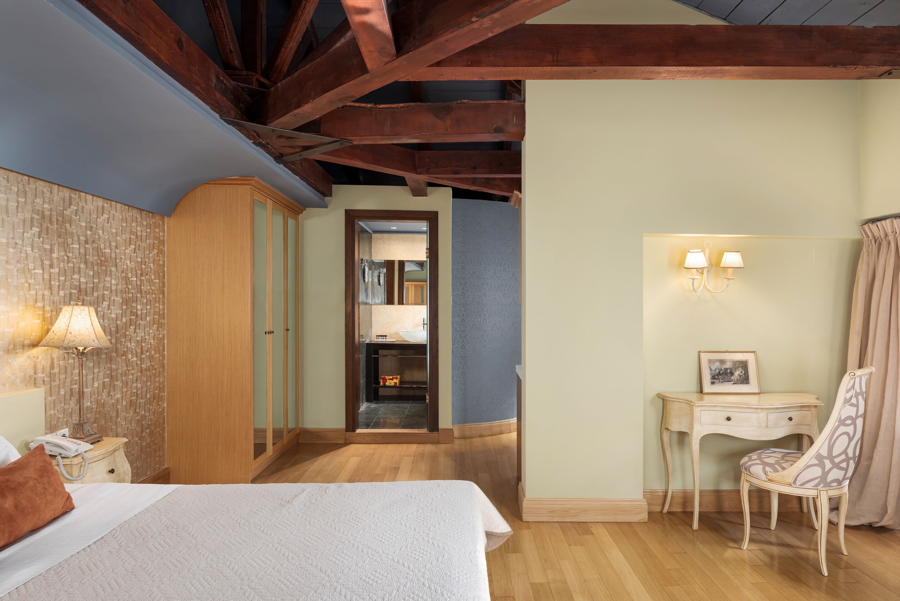 Quarto com cama, mesa de cabeceira, luminária, guarda-roupa, escrivaninha, cadeira e porta que dá para um banheiro.