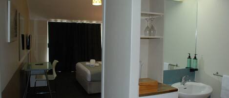 Quarto de hotel com cama, uma pequena mesa com cadeira, mesa de cabeceira com abajur e banheiro com pia e espelho.