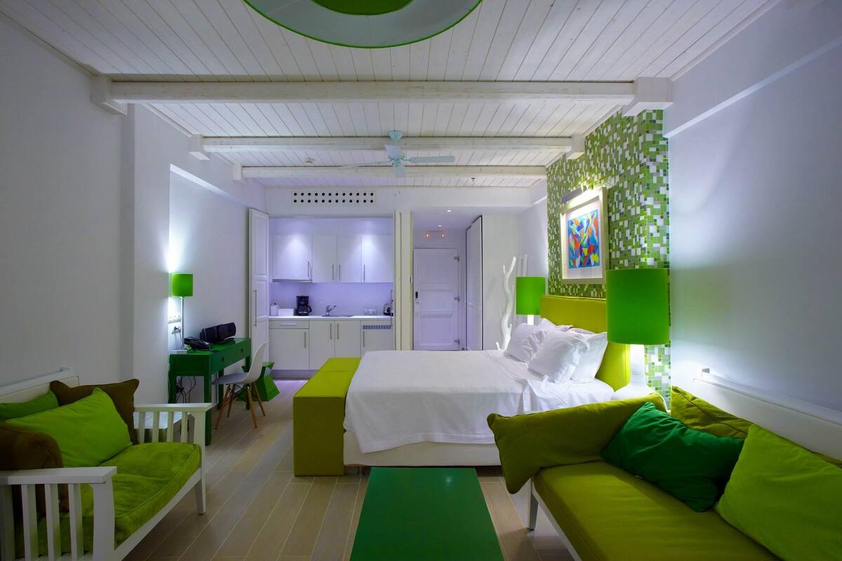 Quarto de hotel moderno com cama, mobiliário verde e branco, escrivaninha e cozinha compacta.