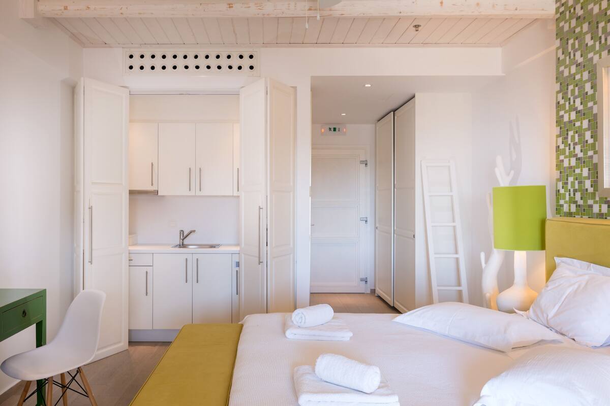 Quarto de hotel com cama, escrivaninha, cadeira, banco verde, pia e espelho.