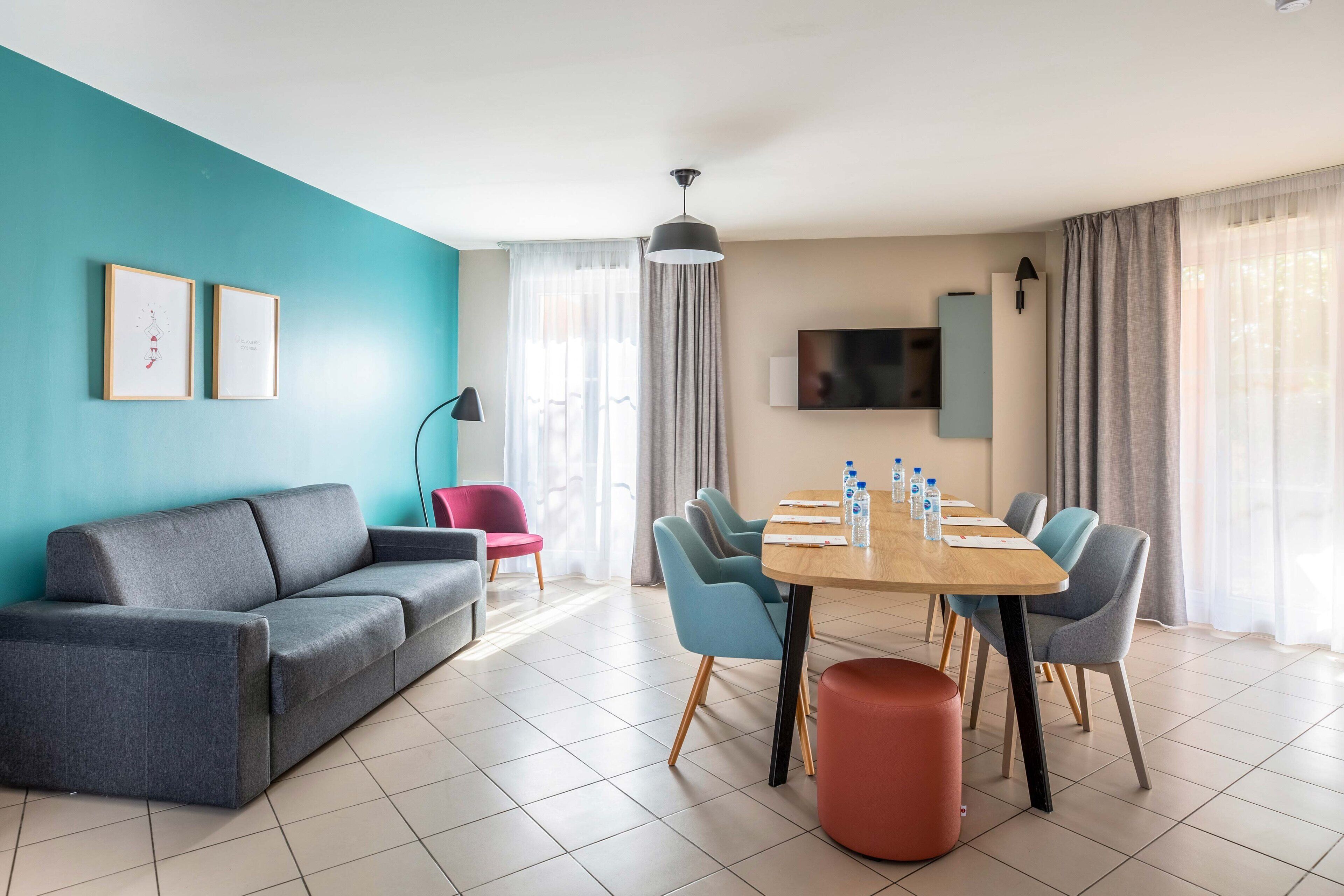 Uma sala de estar moderna com um sofá cinzento, uma mesa de jantar com cadeiras, uma televisão fixada na parede e uma grande janela com cortinas.