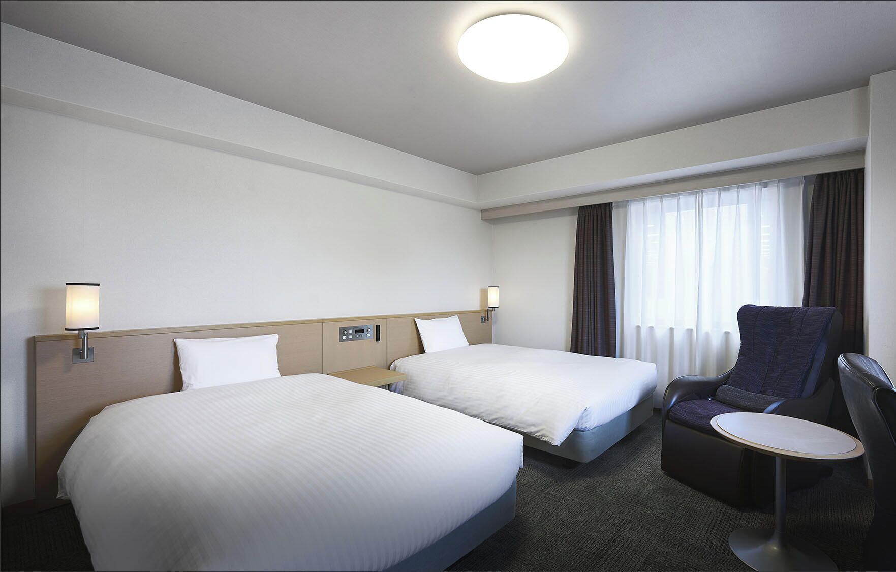 Quarto de hotel com duas camas, uma escrivaninha, uma cadeira e uma mesinha.