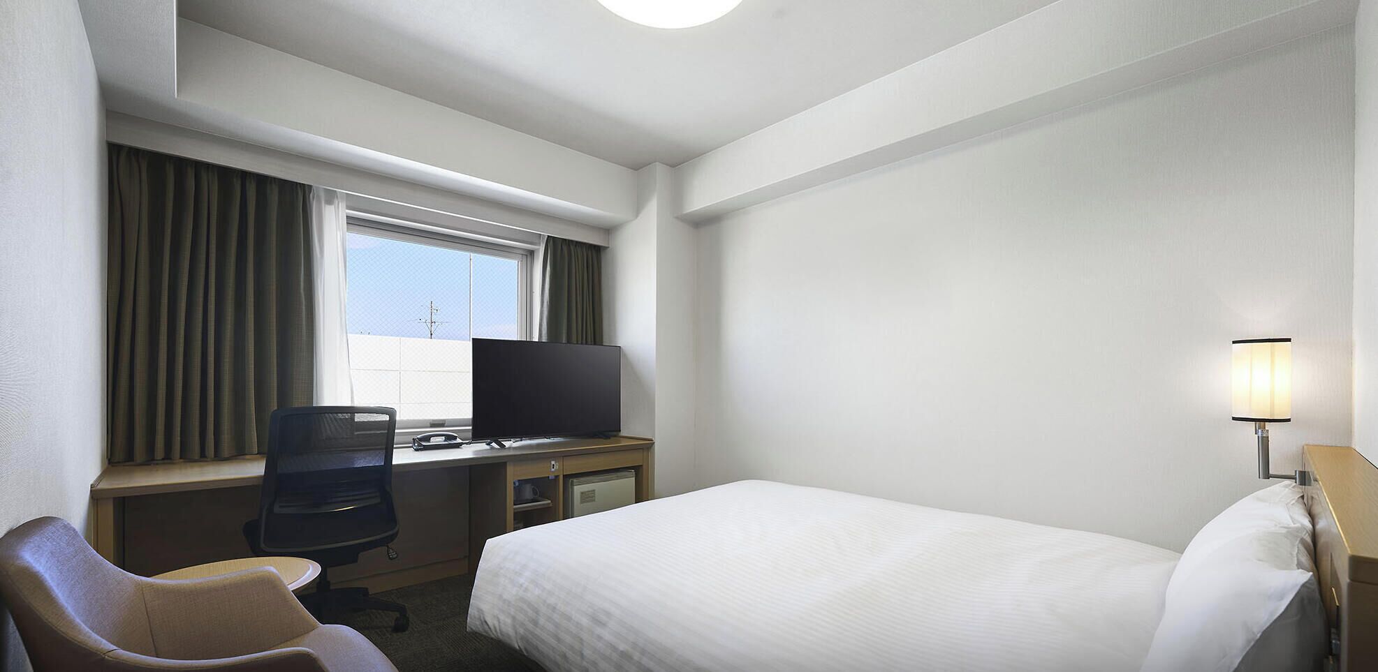 Quarto de hotel com cama, escrivaninha, cadeira, televisão e luminária.