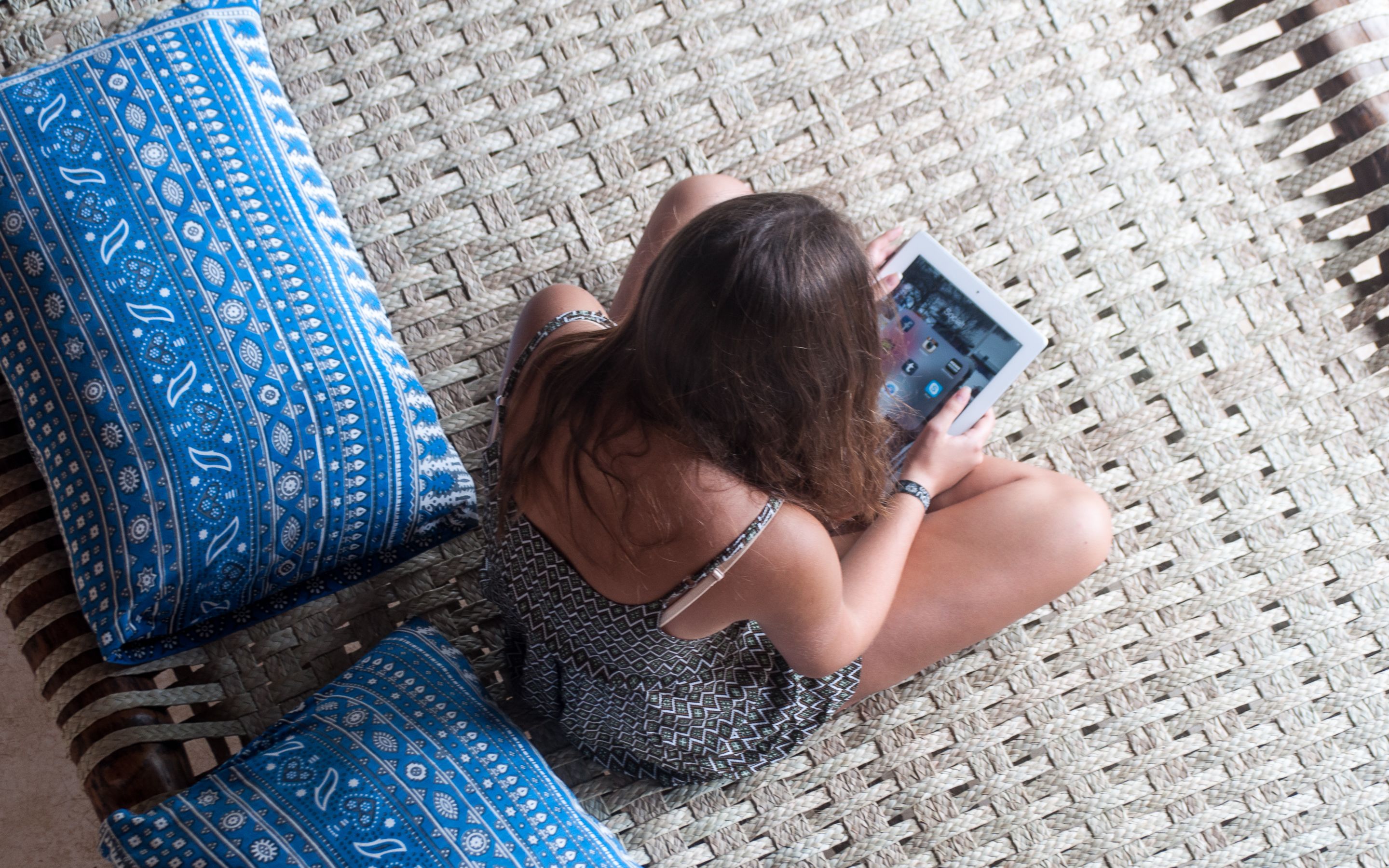 Pessoa sentada em um piso tecido com almofadas azuis estampadas, utilizando um tablet.