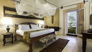 Quarto de hotel com cama, mesas de cabeceira, um banquinho, janela com cortinas e vista para o mar.