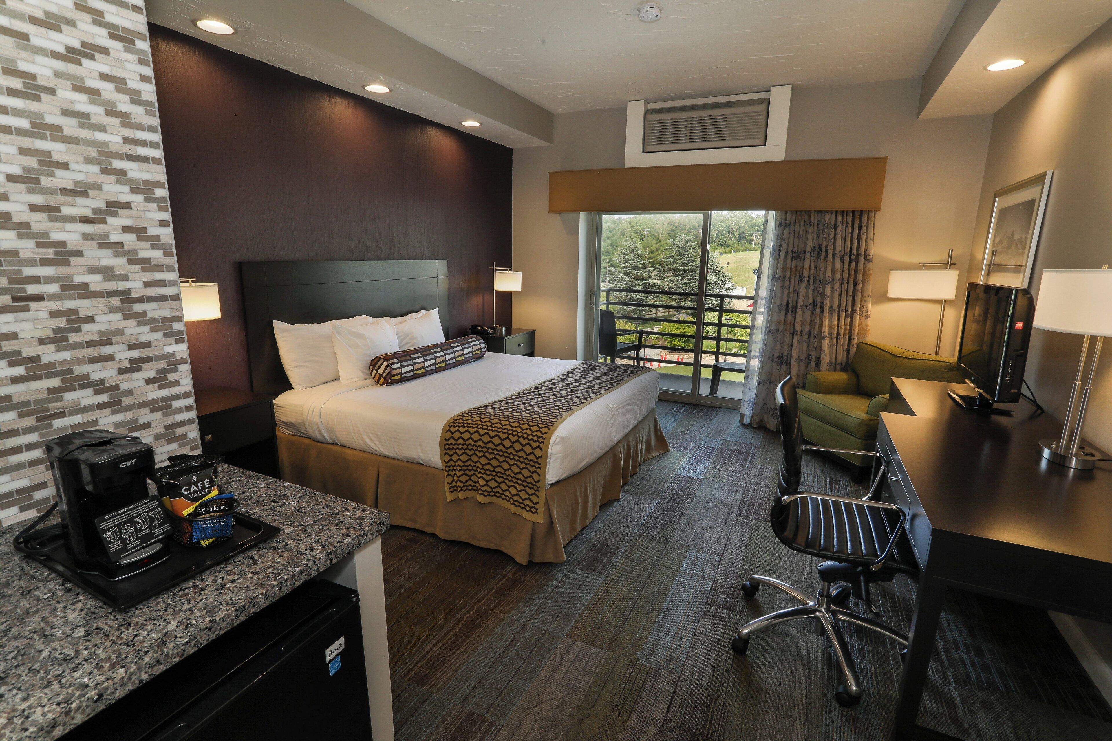 Quarto de hotel com uma cama grande, uma escrivaninha com um computador, uma cadeira e uma varanda com vista para árvores.