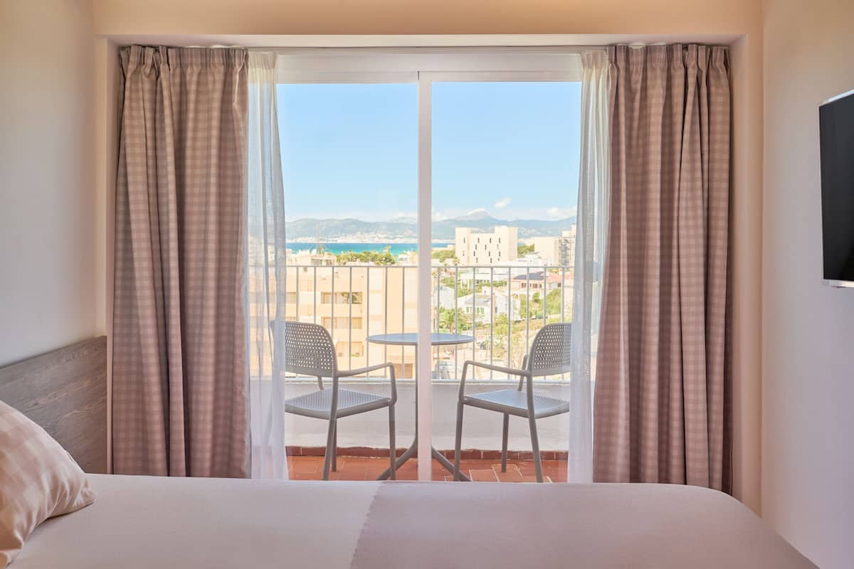 Quarto de hotel com cama, varanda com duas cadeiras e vista para a cidade e o mar.