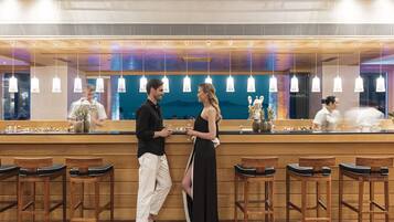 Uma área de bar com banquetas de madeira, um balcão com superfície refletora e luminárias pendentes.