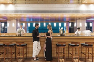 Uma área de bar com banquetas de madeira, um balcão com superfície refletora e luminárias pendentes.