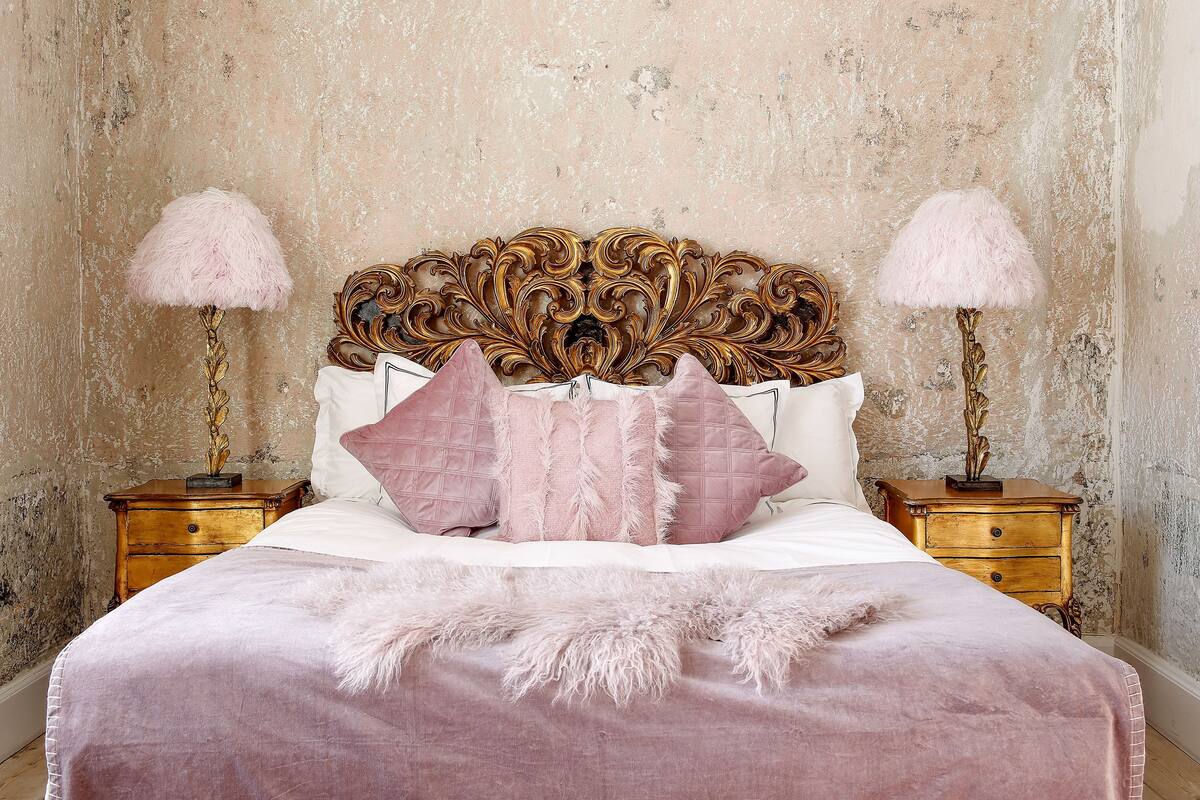 Quarto com uma cama grande, cabeceira ornamentada, duas mesas de cabeceira com abajures e uma parede texturizada.