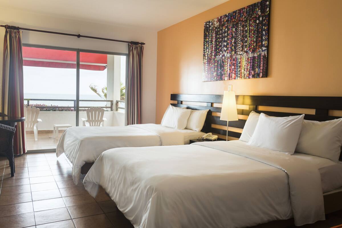 Quarto de hotel com duas camas, uma janela grande com cortinas, uma varanda com cadeiras e uma obra de arte colorida na parede.