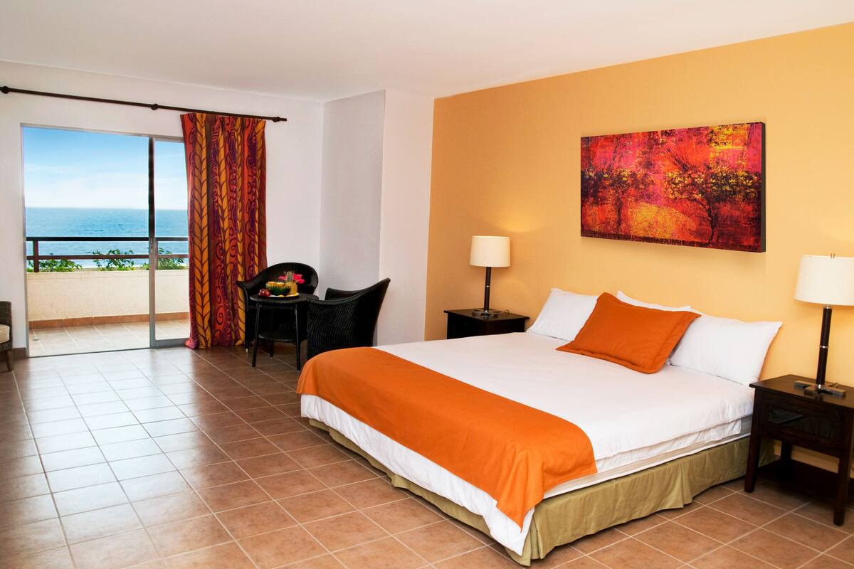 Quarto de hotel com cama, duas mesas de cabeceira com luminárias, uma cadeira, uma mesinha com uma cesta de frutas, varanda com cortinas e um quadro na parede.