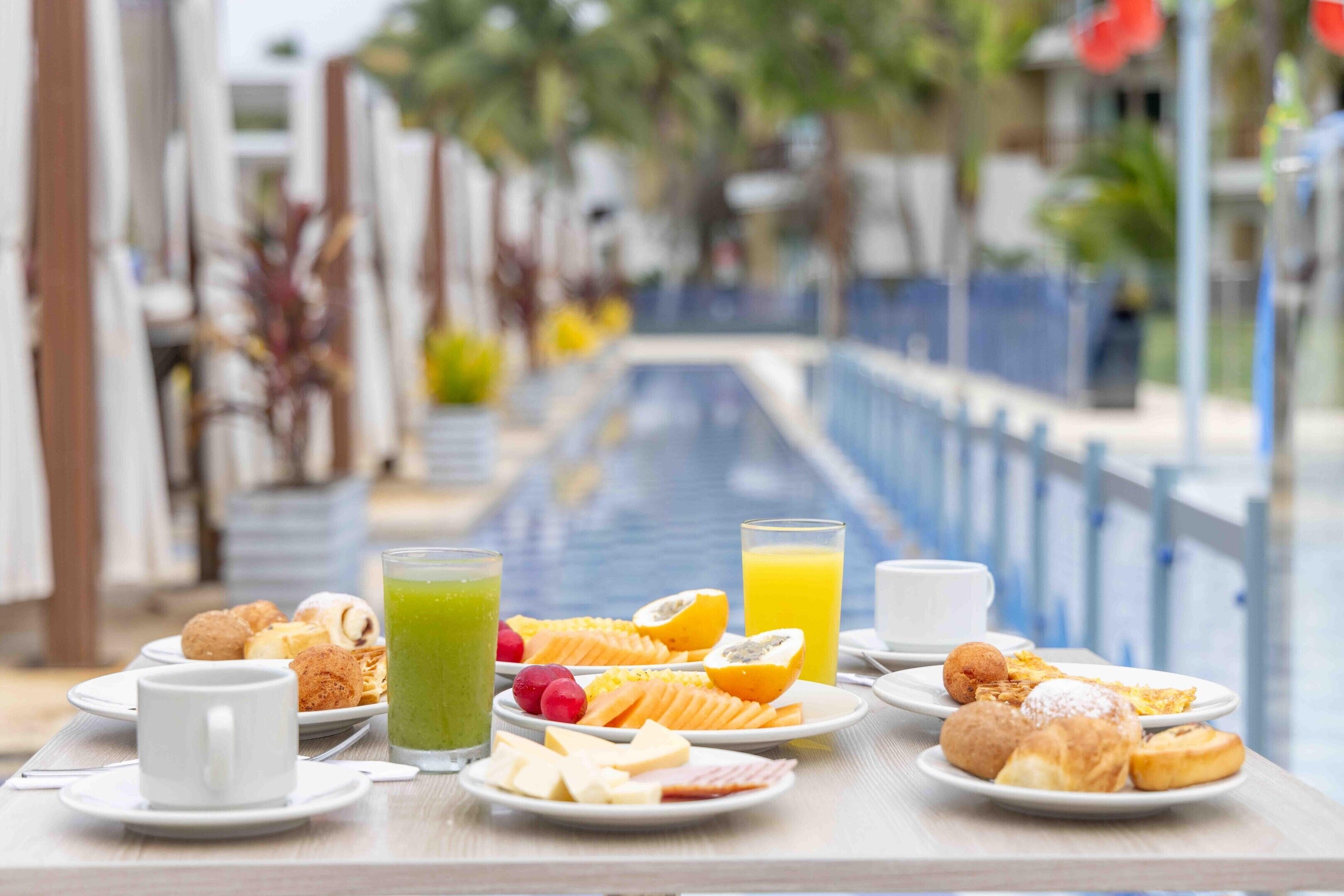 Mesa de café da manhã com diversos alimentos e bebidas, com vista para uma área de piscina.