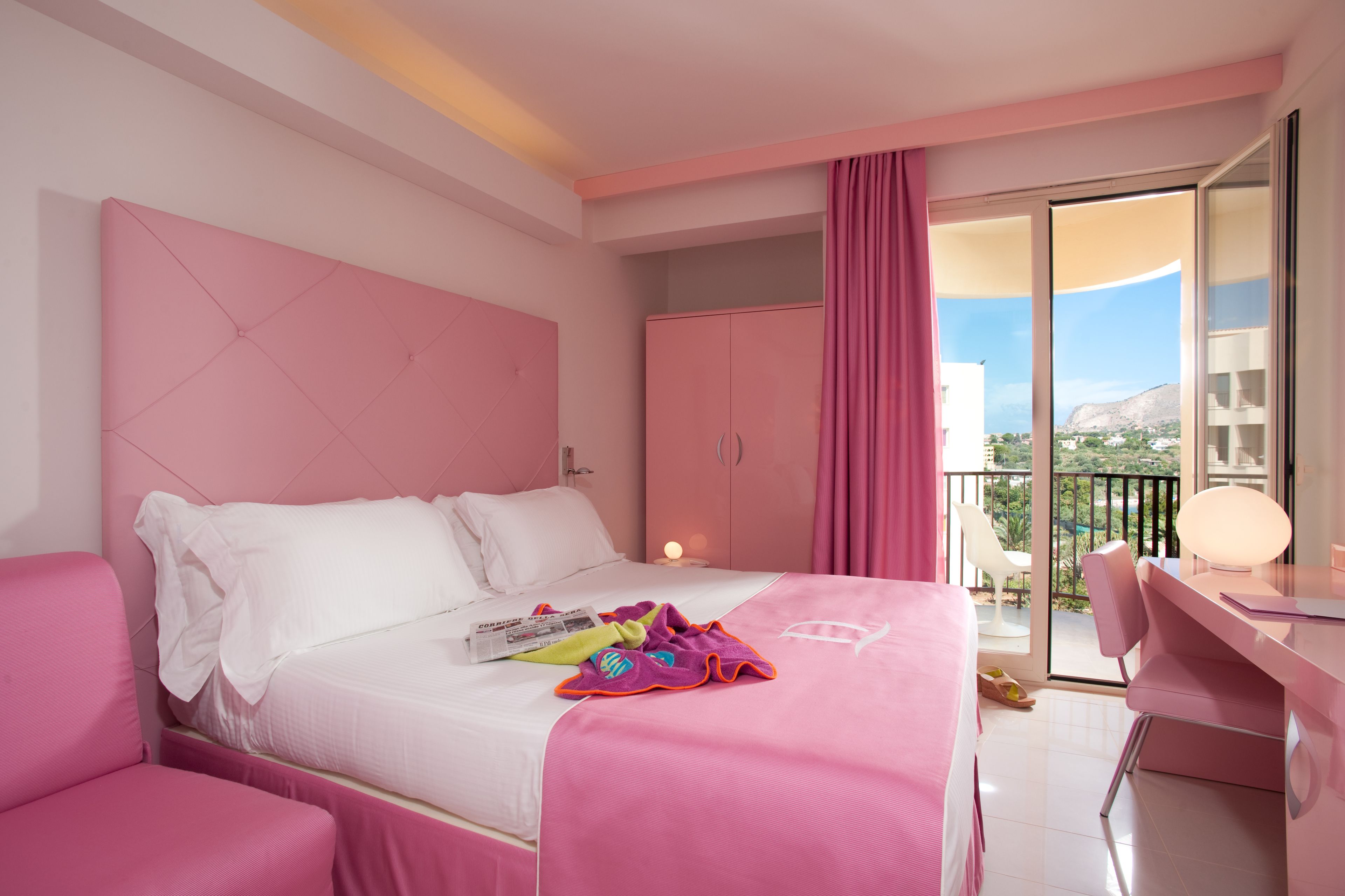 Quarto de hotel em tons de rosa, com cama, escrivaninha, cadeira e varanda com vista.