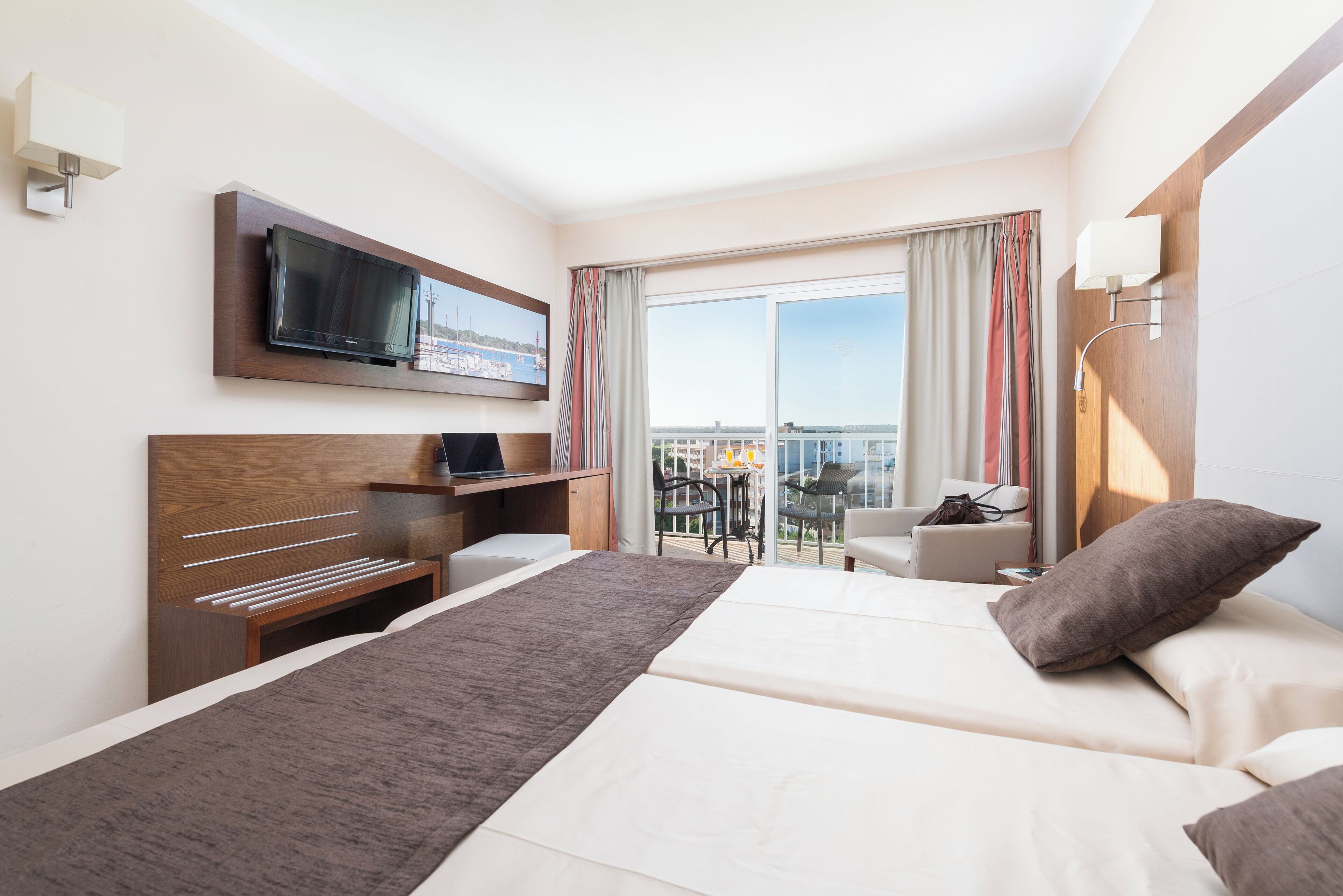 Quarto de hotel com uma cama grande, uma escrivaninha, televisão na parede e varanda com vista para a cidade.