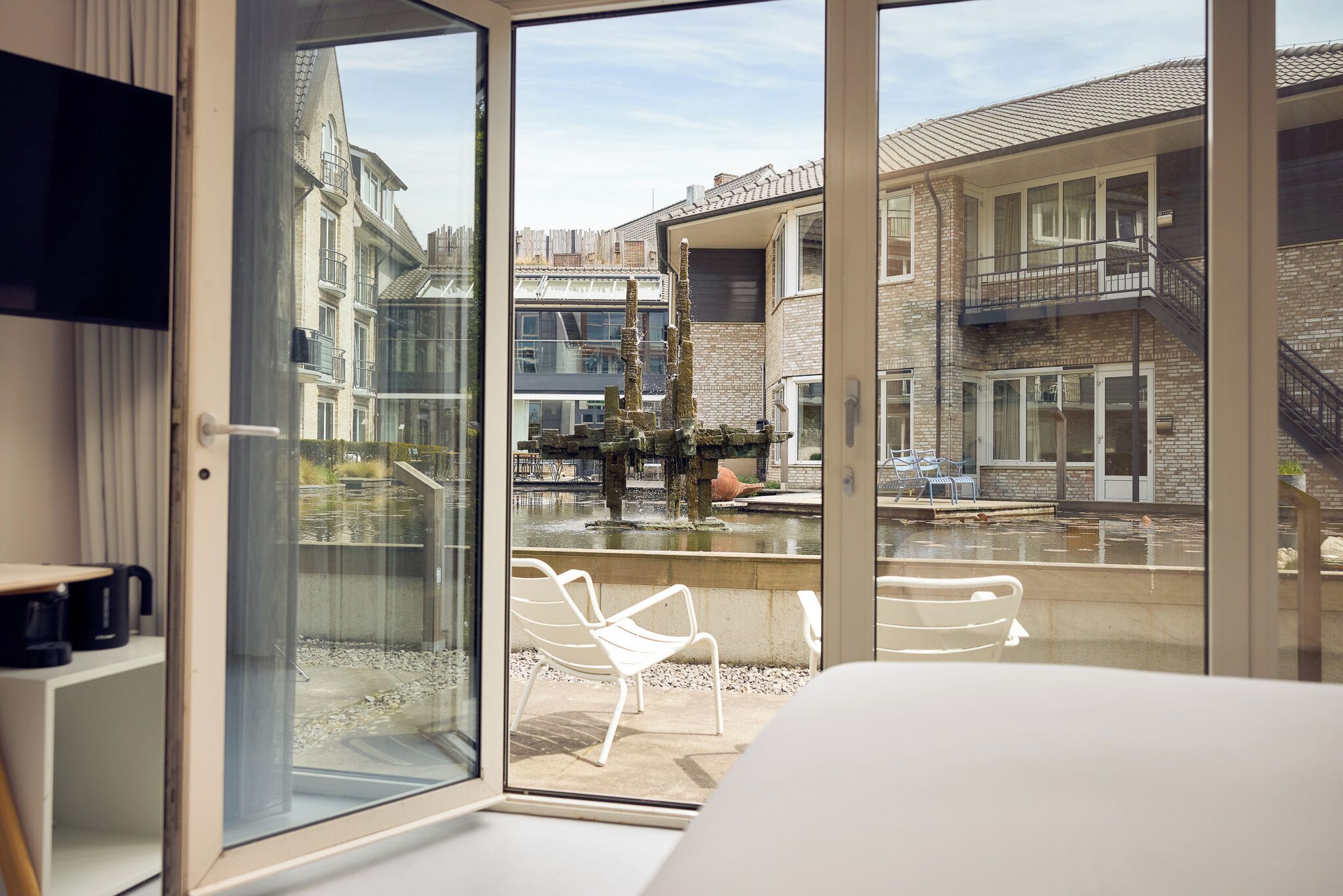 Quarto com uma grande porta de vidro deslizante que dá para um pátio exterior com uma fonte, cadeiras e vista para edifícios vizinhos.