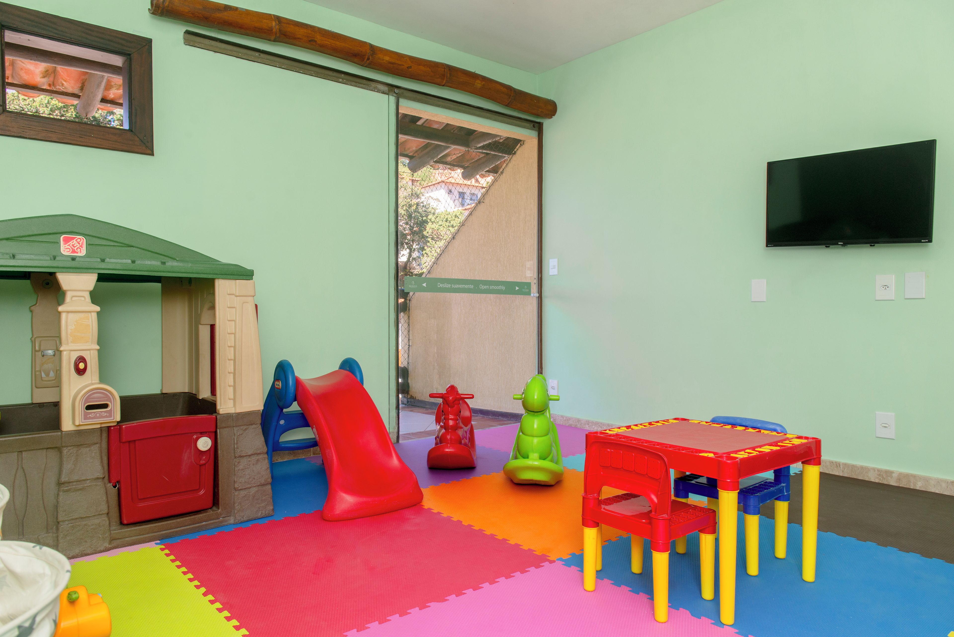 Espaço infantil com tapetes coloridos, escorrega e casinha.